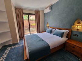 Quarto com cama grande, mesa de cabeceira, luminária na parede e vista para o exterior através de uma porta deslizante.
