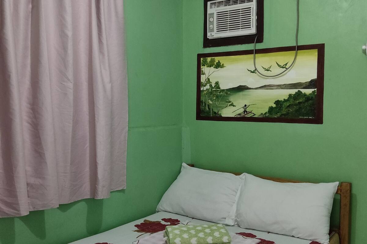 Quarto com cama, paredes verdes, janela com moldura branca, ar-condicionado fixado na parede e pintura de uma paisagem.