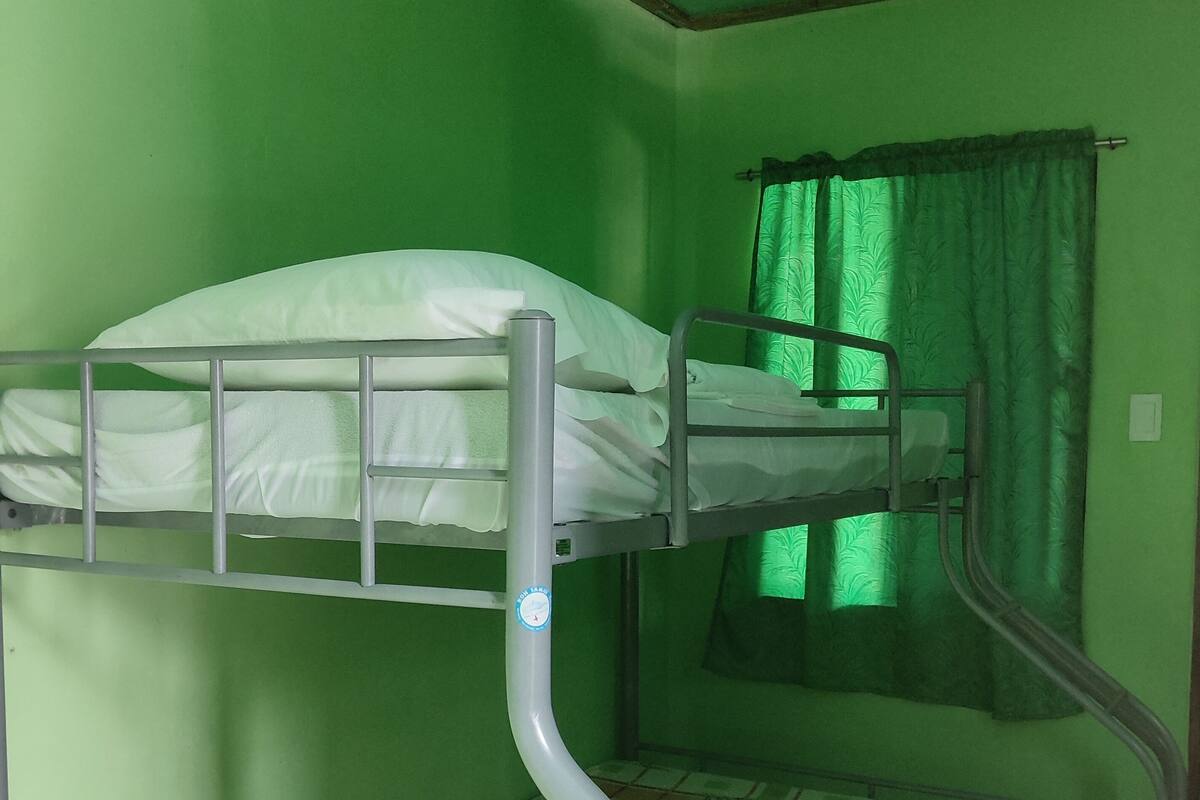 Uma cama de solteiro com colcha xadrez e uma janela com cortinas verdes.