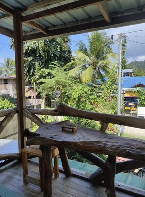 Um deck de madeira com mesa e cadeiras, com vista para um jardim exuberante com palmeiras e um prédio com telhado azul.