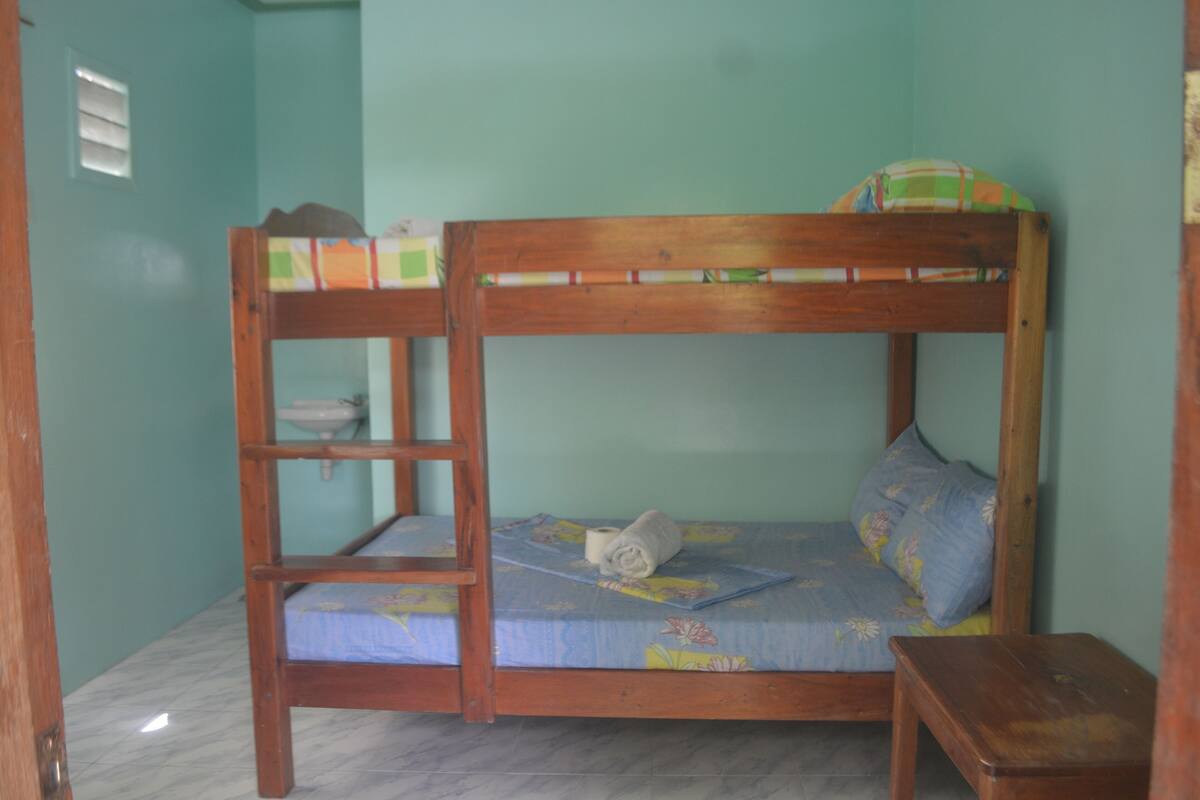 Uma cama beliche com estrutura de madeira e colchão estampado com motivos florais. Um banquinho de madeira pequeno fica ao lado da cama.