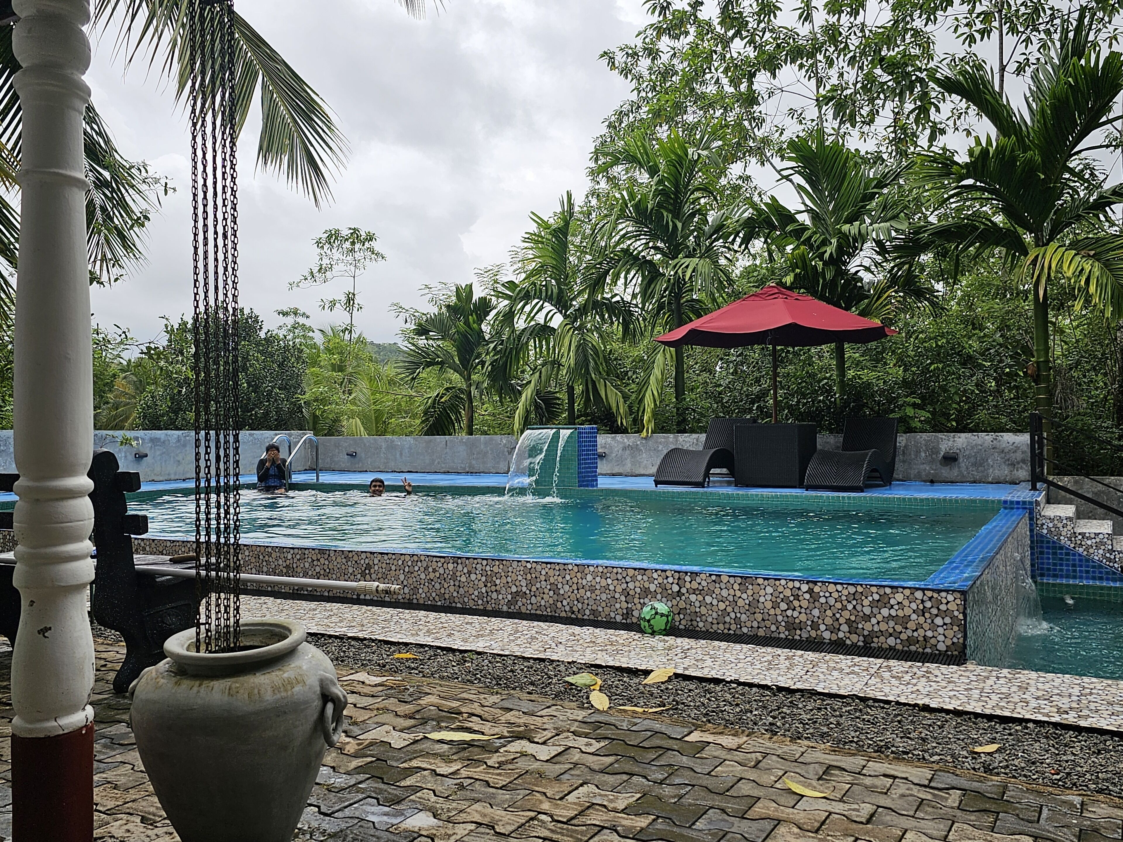 Uma piscina com cascata, rodeada por vegetação exuberante e um guarda-sol vermelho.