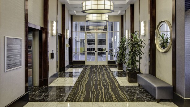 Um lobby de hotel moderno com piso de mármore, um banco, plantas em vasos e uma grande porta de vidro que dá para a área da recepção.