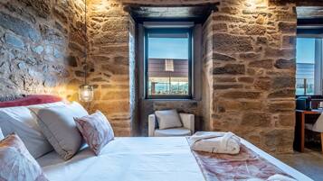 Quarto com paredes de pedra, cama com roupa de cama branca, janela vista para a cidade e uma cadeira pequena.