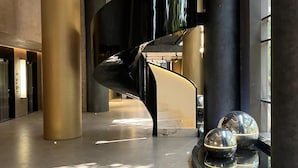 Um interior moderno com uma escada em espiral, colunas cilíndricas imponentes e objetos esféricos refletivos no chão.