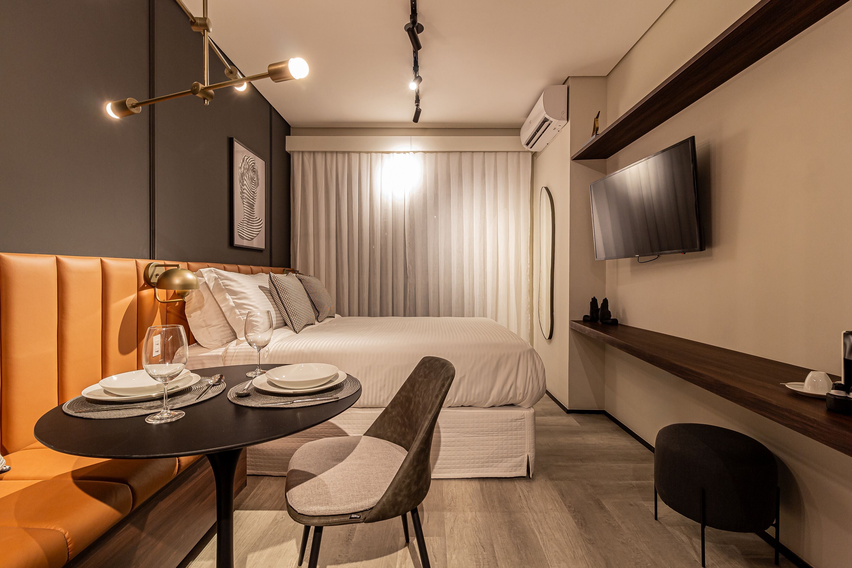 Quarto de hotel moderno com uma cama, uma mesa de jantar com cadeiras, um sofá, uma TV na parede e um ar condicionado.