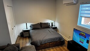 Quarto com cama, mesas de cabeceira, televisão, janela e ar condicionado.