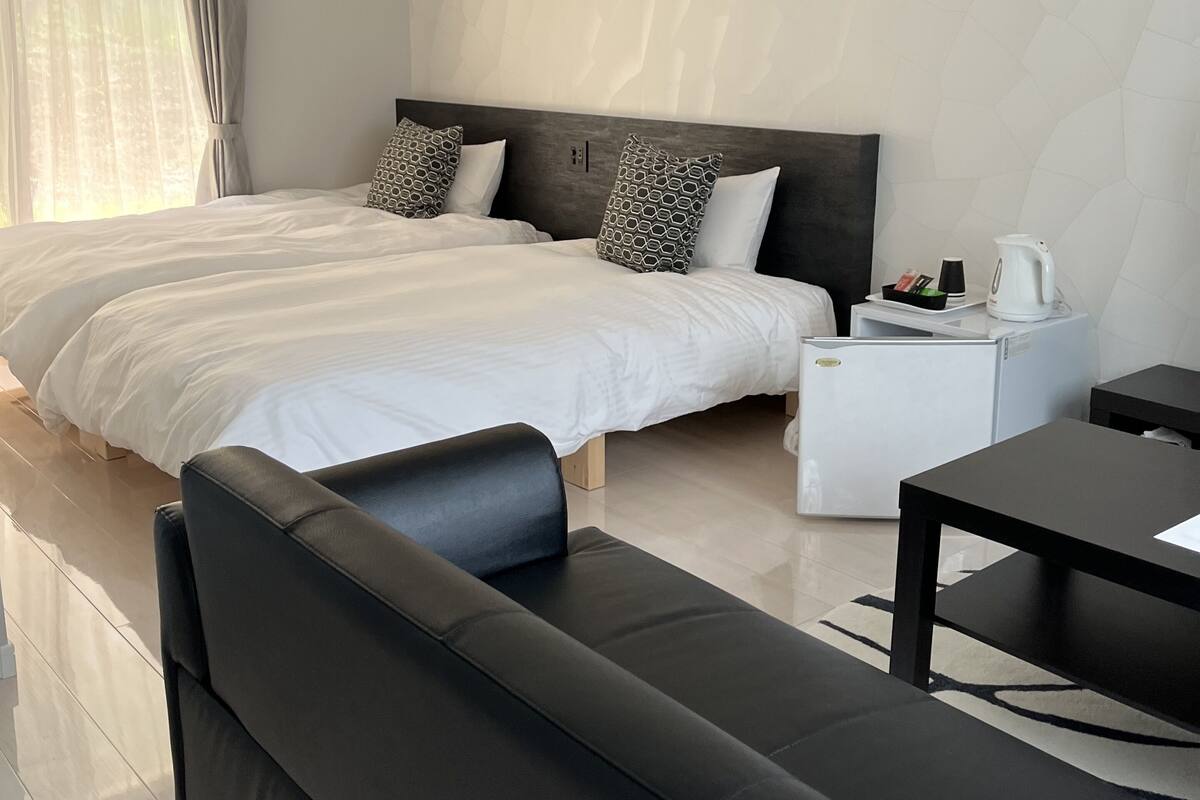Quarto de hotel moderno com uma cama grande, um sofá de couro preto, uma mesa pequena e uma cafeteira branca.