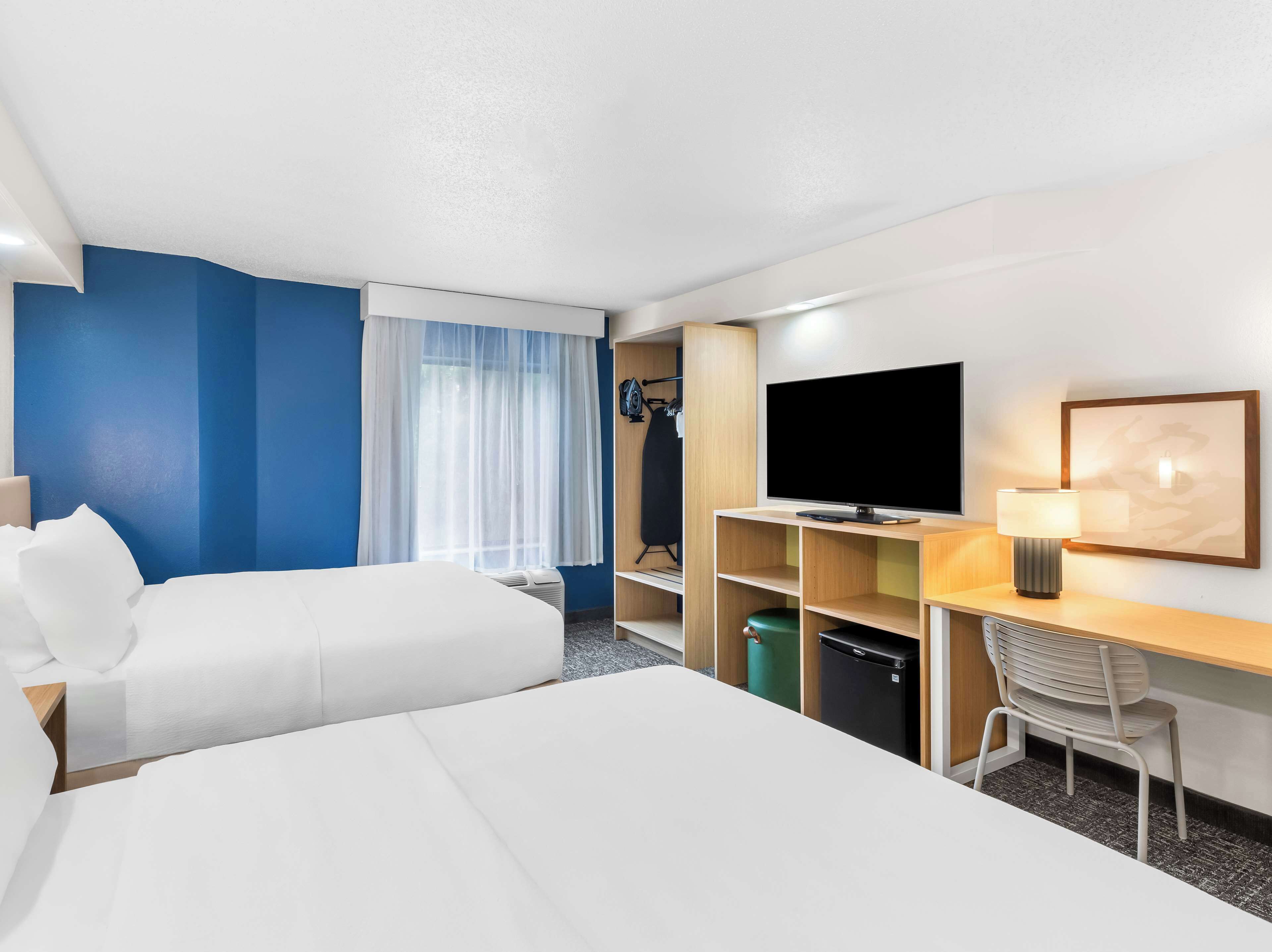 Quarto de hotel com uma cama, escrivaninha com luminária, televisão e armário.