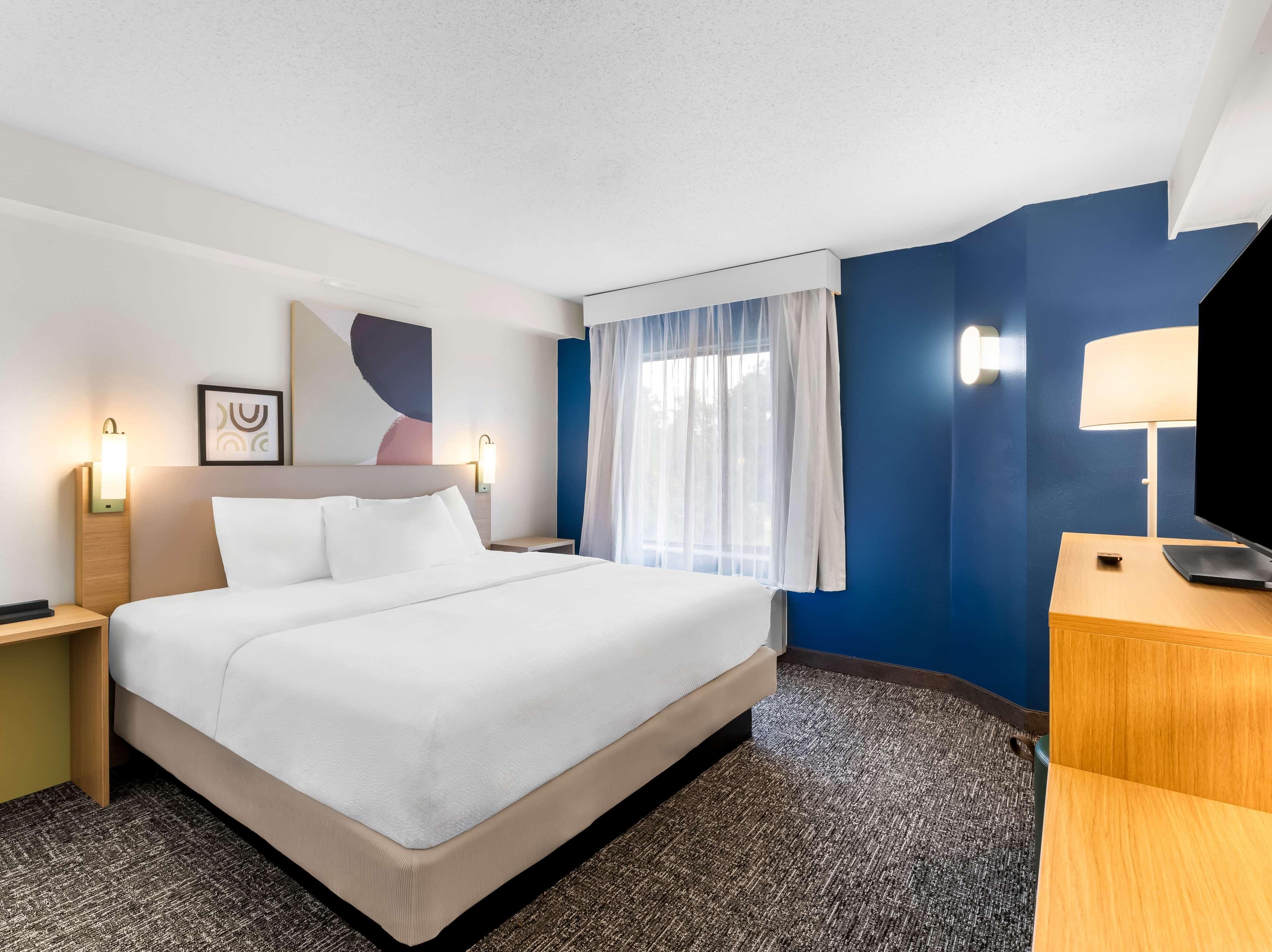 Quarto de hotel com cama, mesas de cabeceira, abajur, televisão e janela com cortinas.