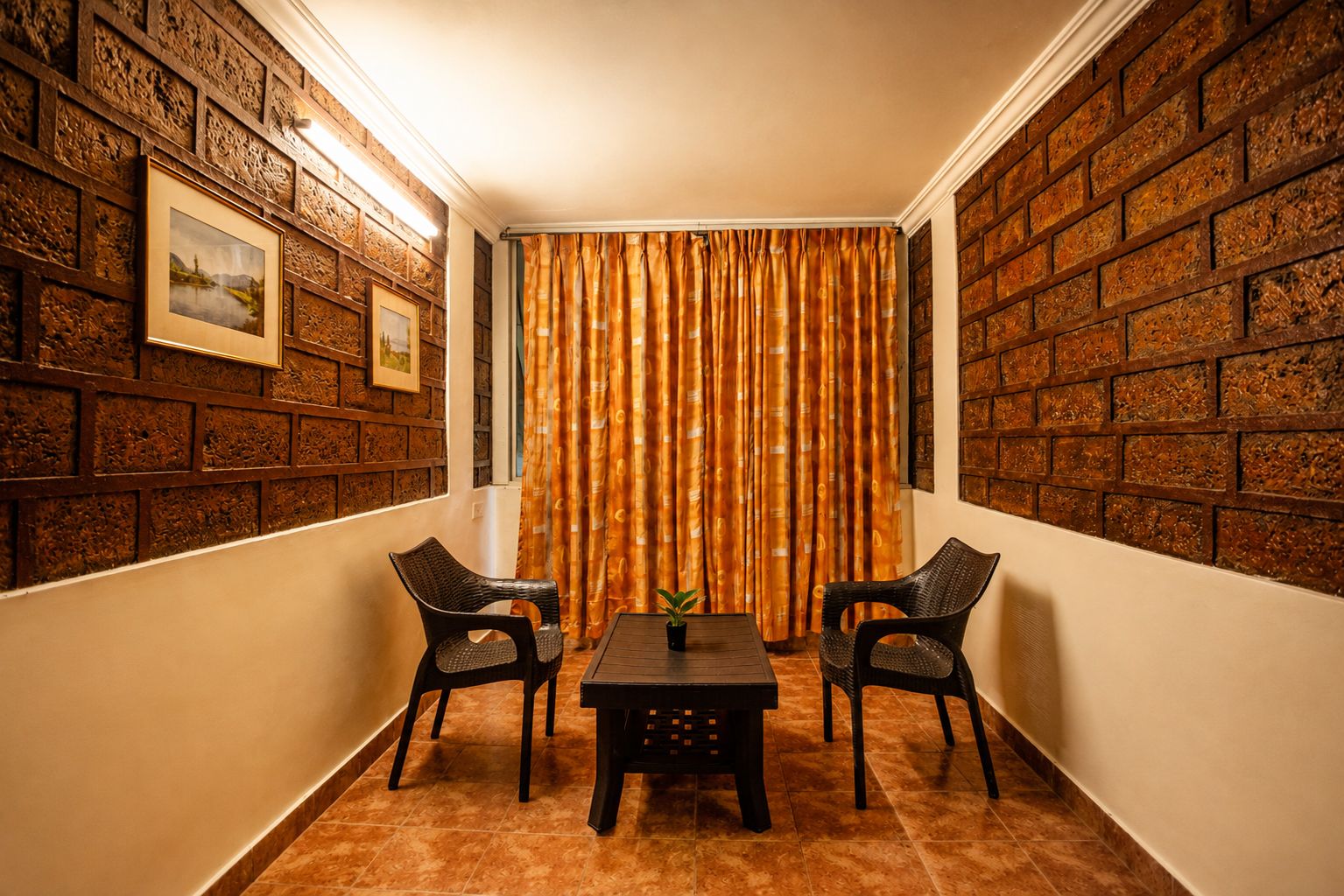Um quarto pequeno com parede de tijolos, duas cadeiras de vime, uma mesinha e uma cortina.