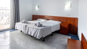 Quarto de hotel com cama de casal, duas mesas de cabeceira, uma mesinha de madeira e uma janela grande com vista para edifícios.