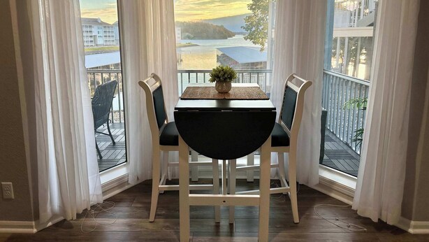 Uma sala de jantar com mesa e cadeiras, uma planta em vaso e vista para um lago e edifícios.