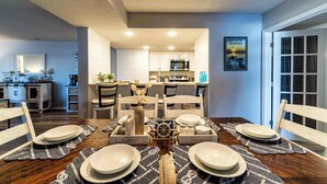 Uma sala de jantar com uma mesa de madeira posta para quatro pessoas, rodeada por cadeiras brancas. Uma cozinha é visível ao fundo, com armários brancos e eletrodomésticos de aço inoxidável.