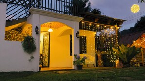 Uma casa com fachada branca, uma parede de pedra e cerca de madeira. Há luzes de Natal e um vaso com planta perto da entrada.