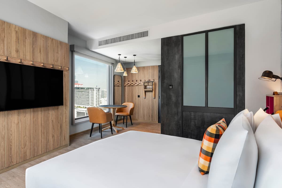 Quarto de hotel moderno com uma cama grande, TV fixada na parede, área de refeições com cadeiras e vista da cidade através de janelas amplas.