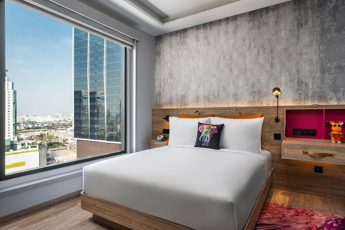 Quarto de hotel moderno com uma cama grande, vista para a cidade, parede de destaque em concreto e mesa de cabeceira com abajur.
