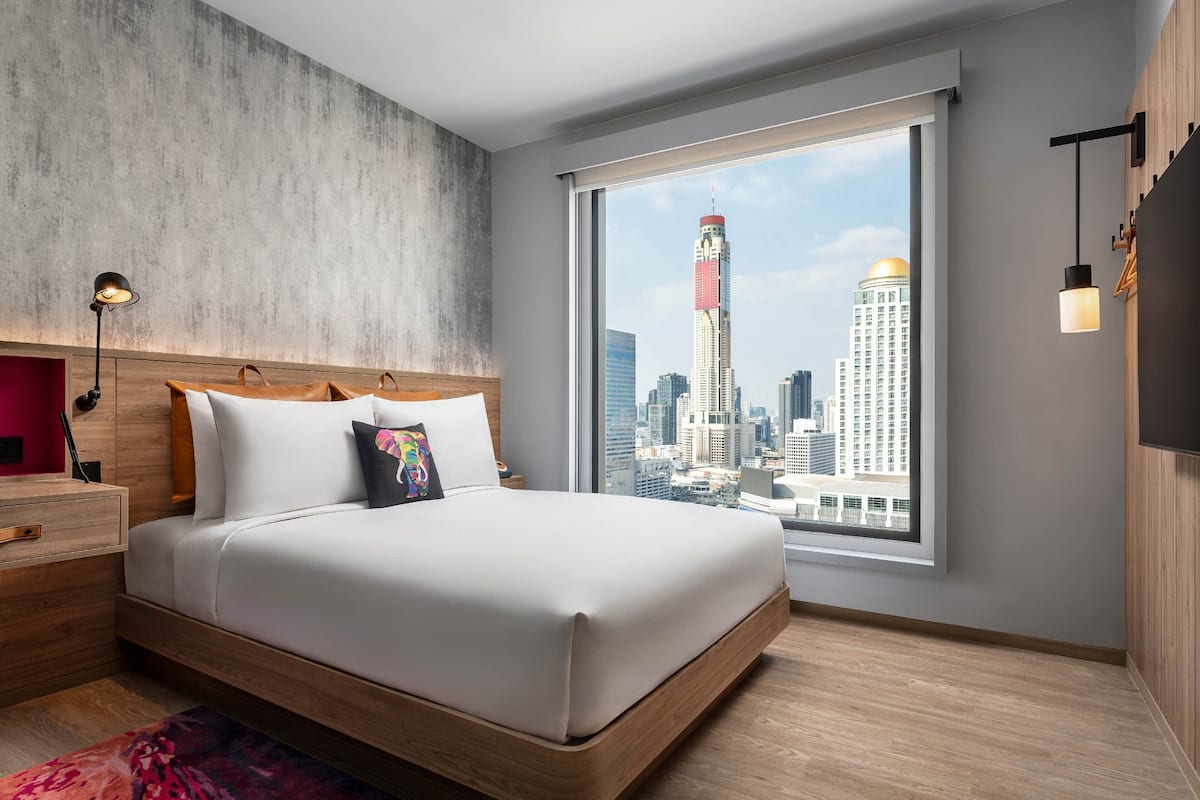 Quarto de hotel moderno com uma cama grande, vista para a cidade, cabeceira de madeira e mesa de cabeceira com uma luminária.