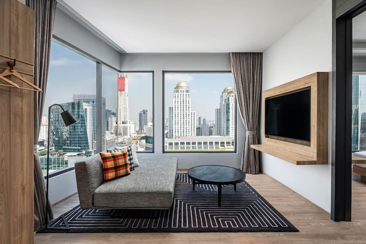 Quarto de hotel moderno com uma grande janela oferecendo vista para a cidade, um sofá com manta estampada, uma pequena mesa redonda e uma televisão na parede.