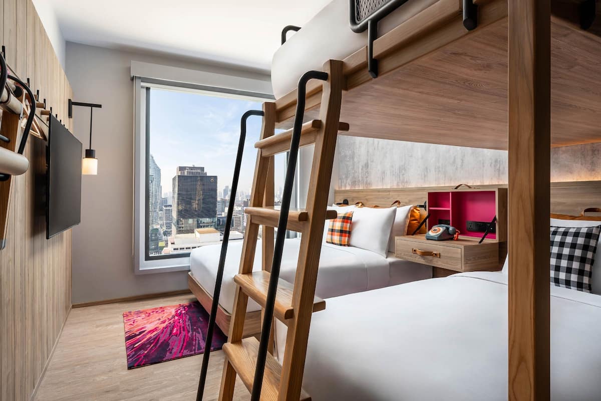 Quarto de hotel moderno com beliche, vista para a cidade, escada de madeira, escrivaninha com telefone e prateleira rosa.