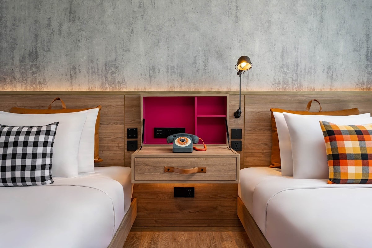 Quarto de hotel com duas camas, uma cabeceira de madeira, uma prateleira rosa, um telefone e um abajur.