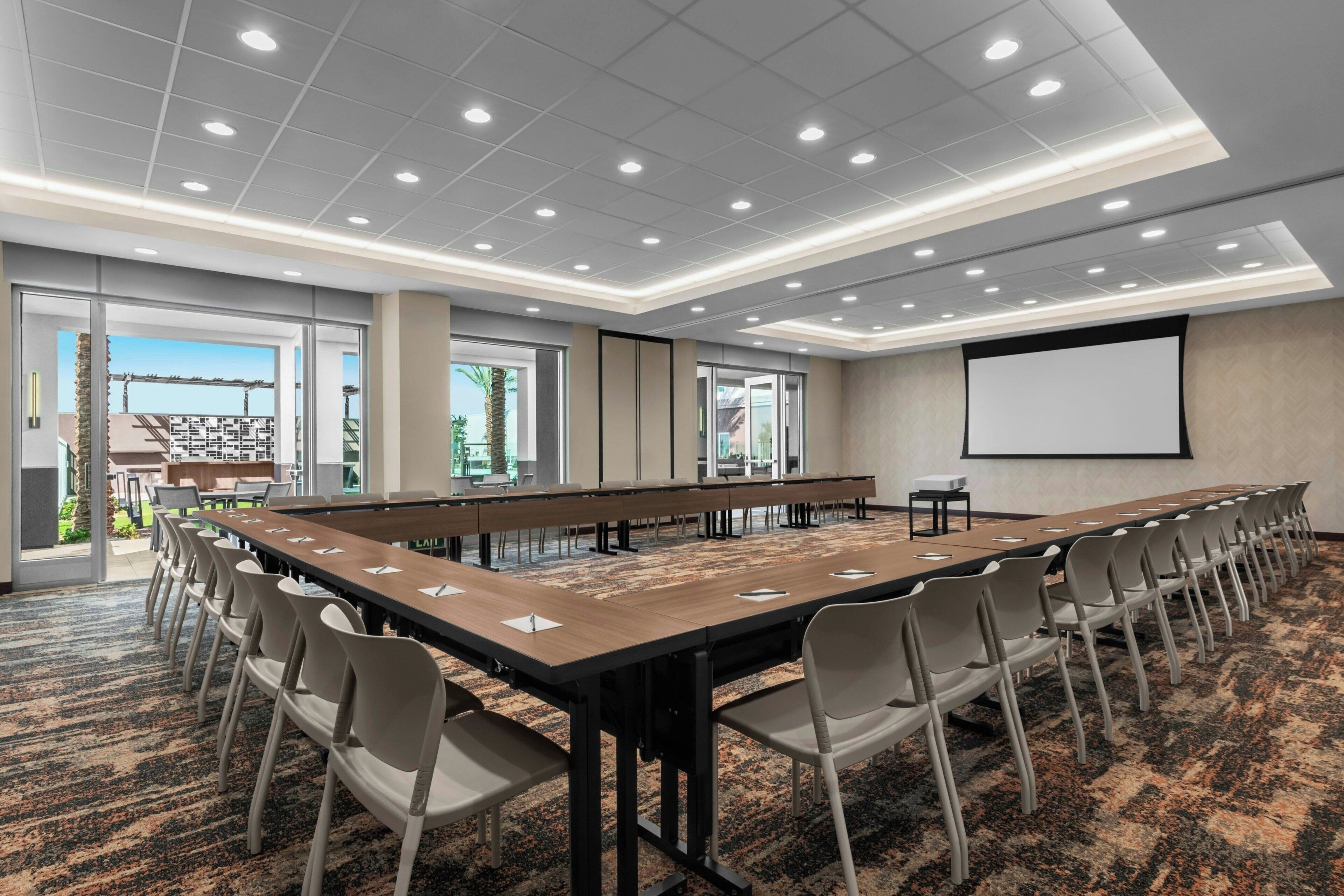 Uma sala de conferência com uma mesa comprida, cadeiras, tela de projetor e amplas janelas.