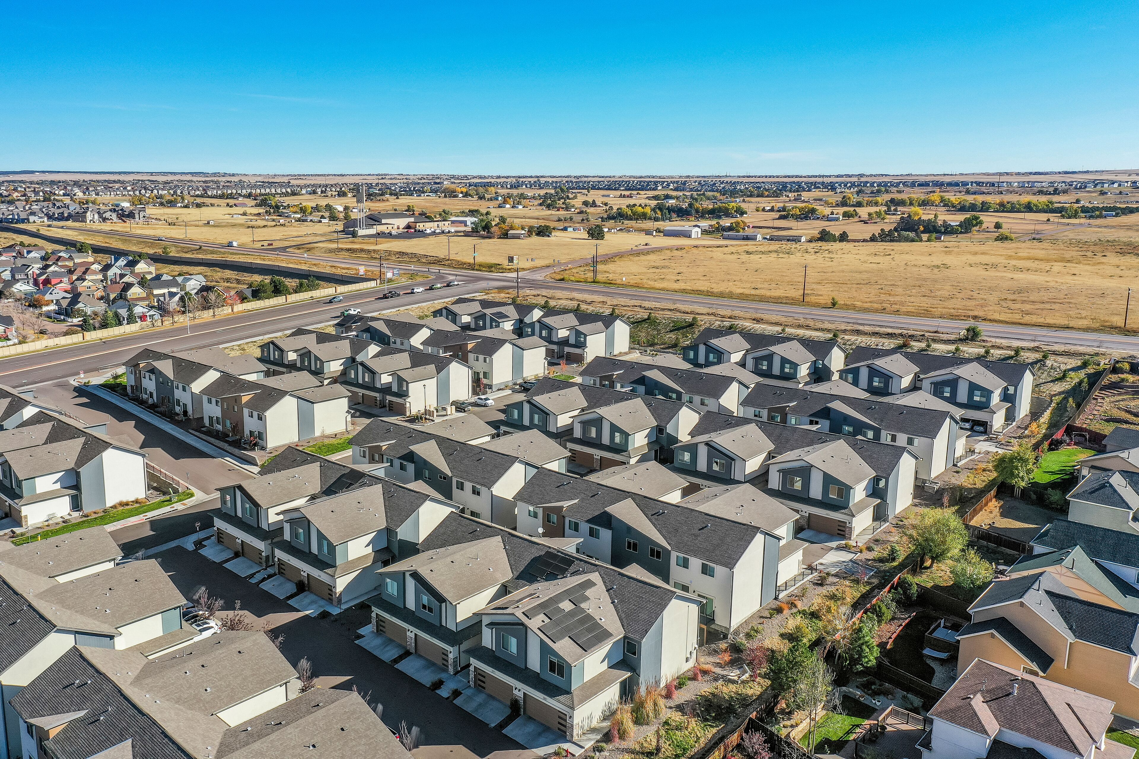 Zona residencial com casas modernas dispostas em um padrão de grade, apresentando uma combinação de construções térreas e com dois andares, com uma rua visível em primeiro plano e campos abertos ao fundo.