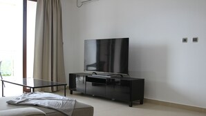 Uma sala de estar moderna com uma televisão de tela plana sobre um móvel preto, um sofá bege e uma mesa de centro de vidro.