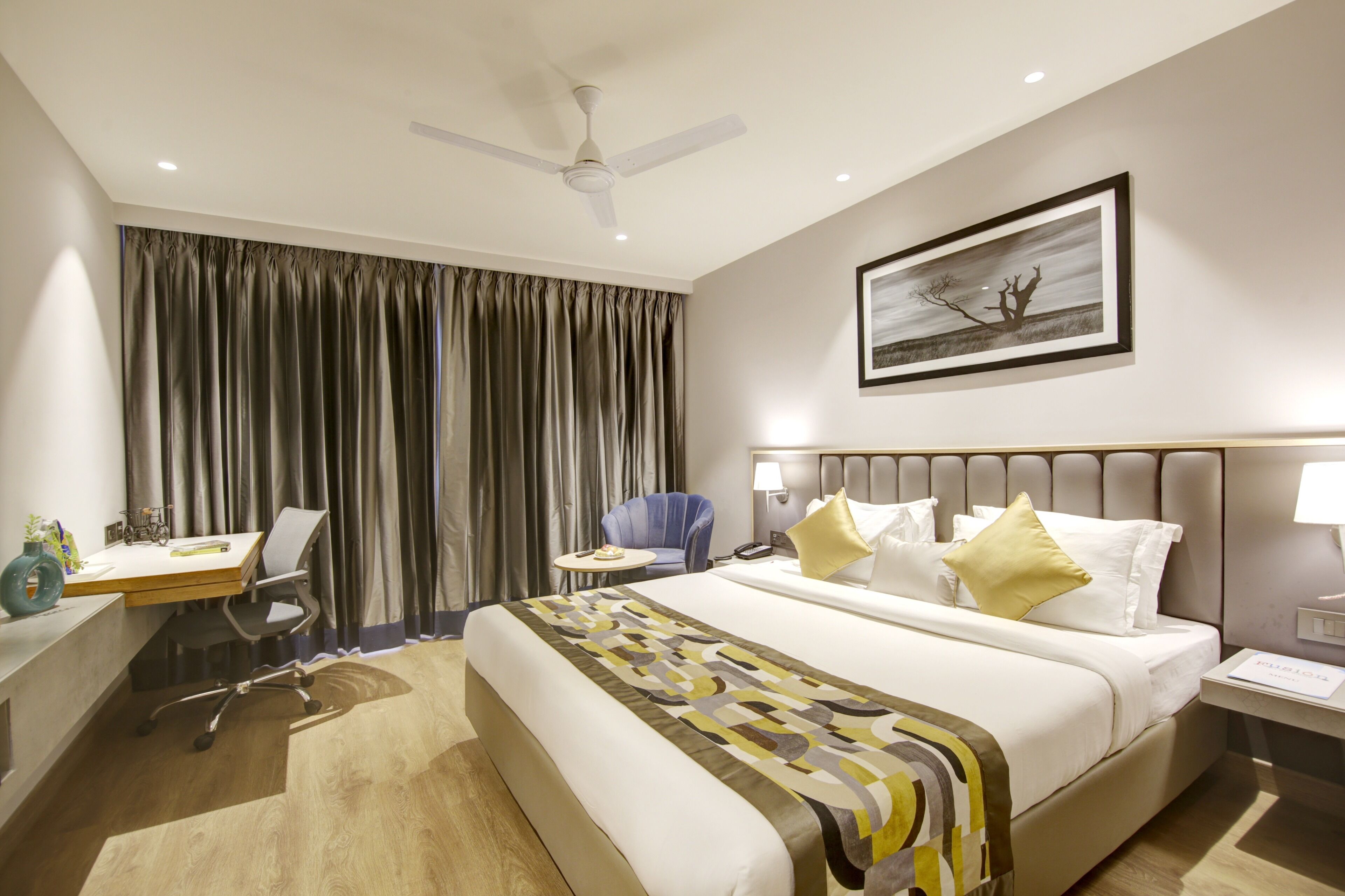 Quarto de hotel com cama, escrivaninha, cadeira e ventoinha no teto.