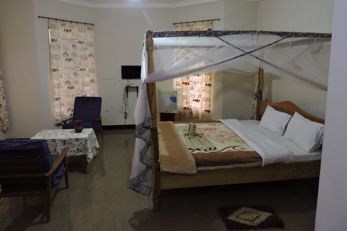 Quarto de hotel com cama de dossel, uma mesa pequena com toalha de mesa florida, uma cadeira e uma televisão na parede.