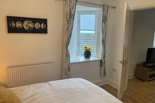 Quarto com cama, radiador, janela com cortinas, vaso com flores e porta que dá para outro cômodo.