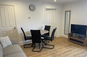 Uma sala de estar moderna com uma mesa de jantar redonda, cadeiras pretas, uma televisão de tela plana sobre um suporte de madeira e um sofá cinza.