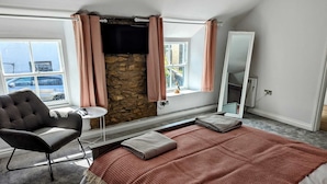 Quarto com cama, uma cadeira, mesinha, parede de pedra, janela e espelho.