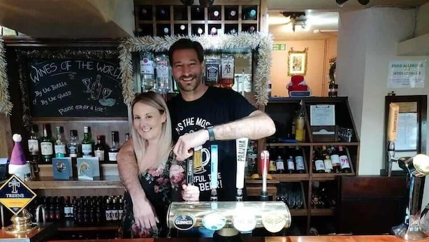 Um bar com menu negro exibindo vinhos e cervejas, um bartender e uma mulher atrás do balcão.