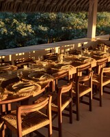 Uma área de refeições ao ar livre com mesas e cadeiras de madeira, disposta com louça fina, copos e velas.