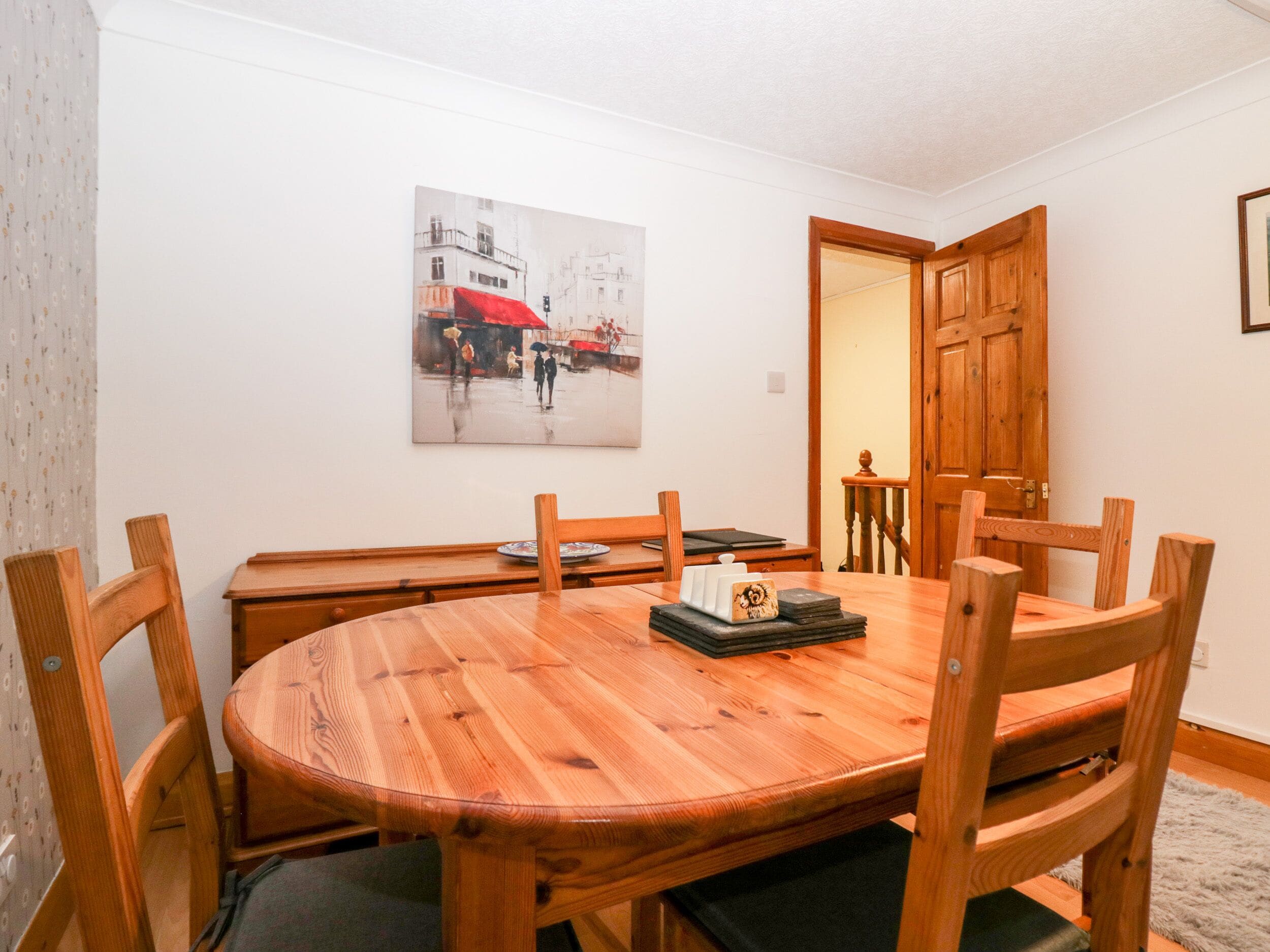 Uma mesa de jantar de madeira com seis cadeiras, um quadro na parede e uma porta de madeira ao fundo.