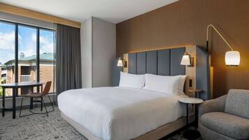 Quarto de hotel com uma cama grande, uma mesa, uma cadeira e um sofá. Há duas luminárias fixadas na parede e vista para o exterior através de amplas janelas.