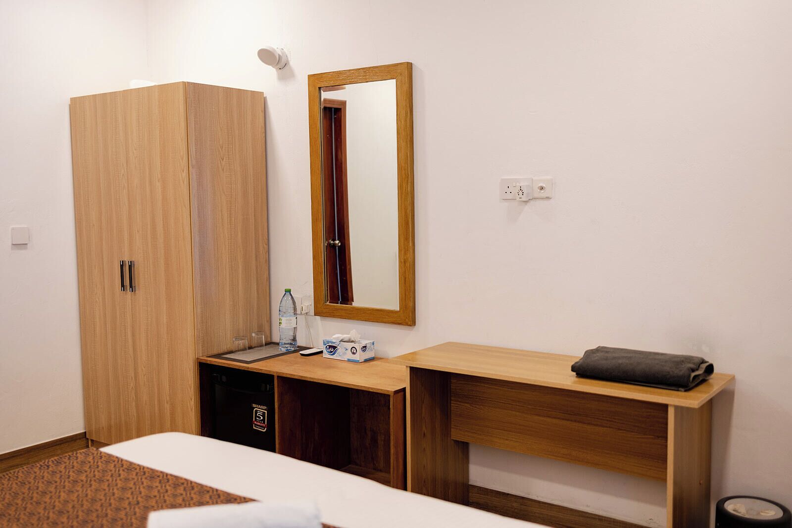 Quarto de hotel com guarda-roupa de madeira, uma secretária pequena com uma toalha, um espelho e uma garrafa de água.