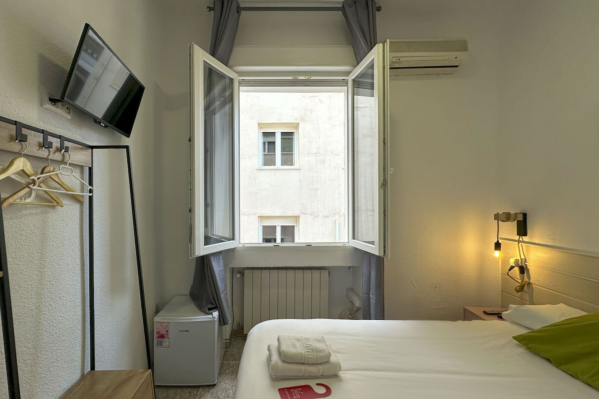 Quarto de hotel com cama, televisão na parede, radiador e janela com cortinas.
