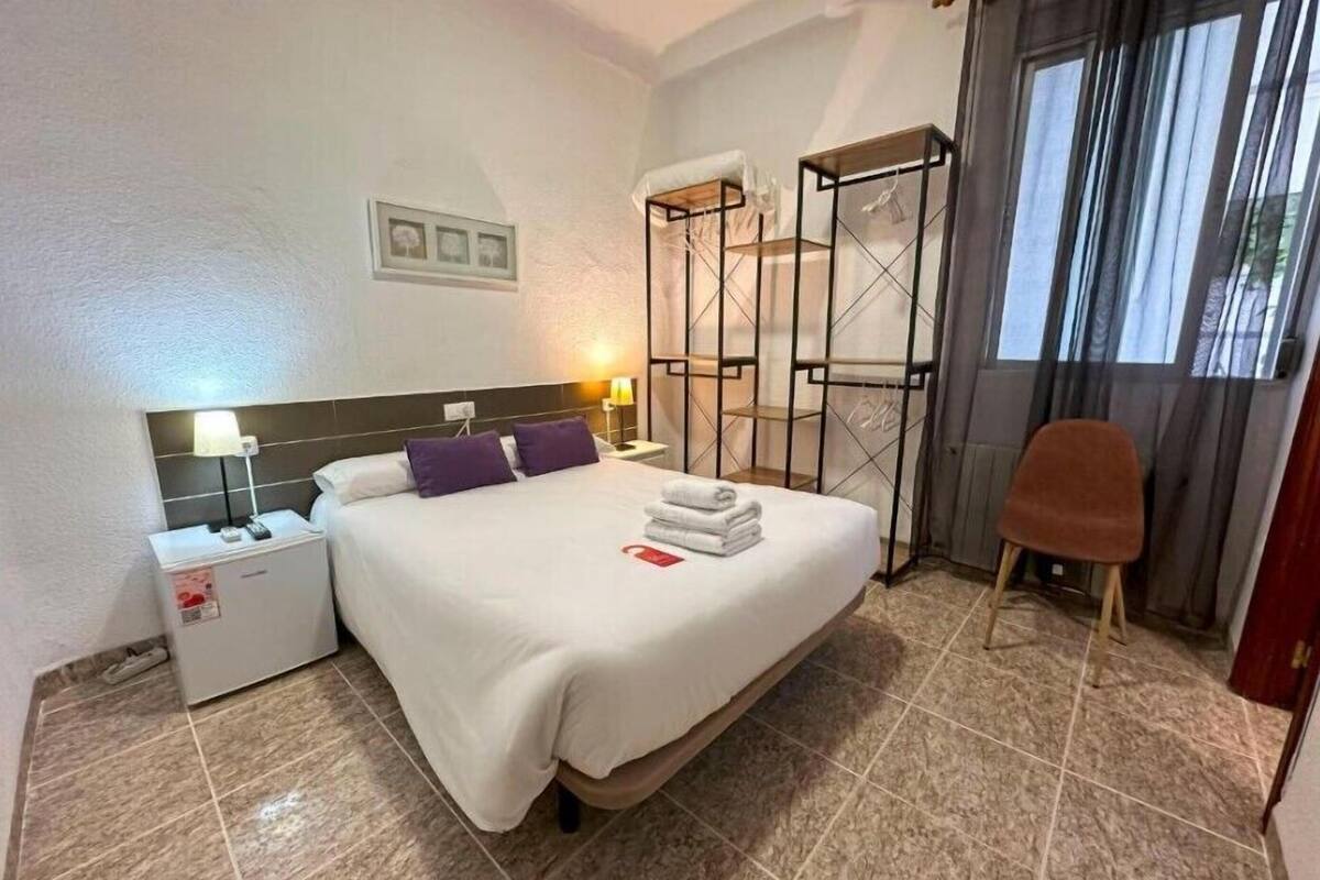 Quarto de hotel com cama, mesa de cabeceira, cadeira, janela com cortinas e prateleira fixada na parede.