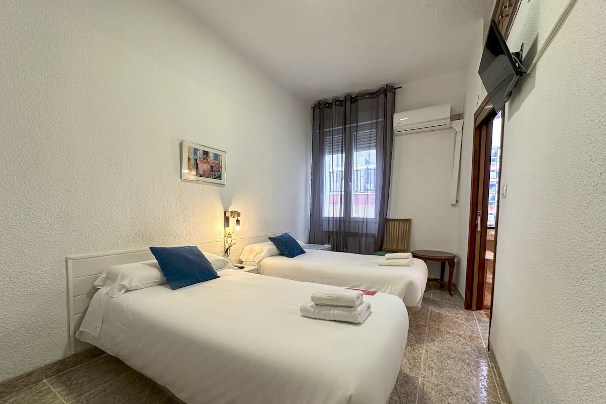 Quarto de hotel com duas camas, uma mesa pequena, uma cadeira, televisão presa à parede e janela com cortinas.