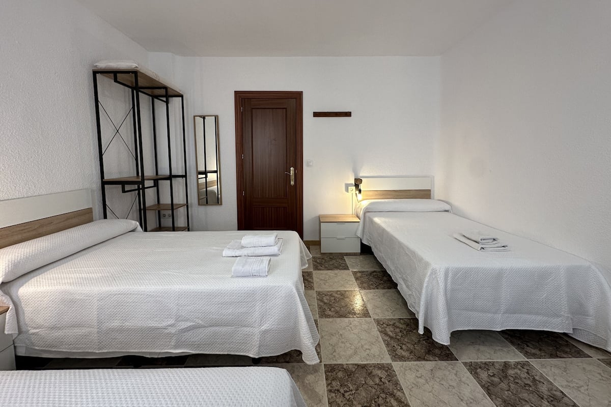 Quarto de hotel com duas camas de solteiro, uma porta de madeira, uma mesa de cabeceira com abajur e um espelho na parede.