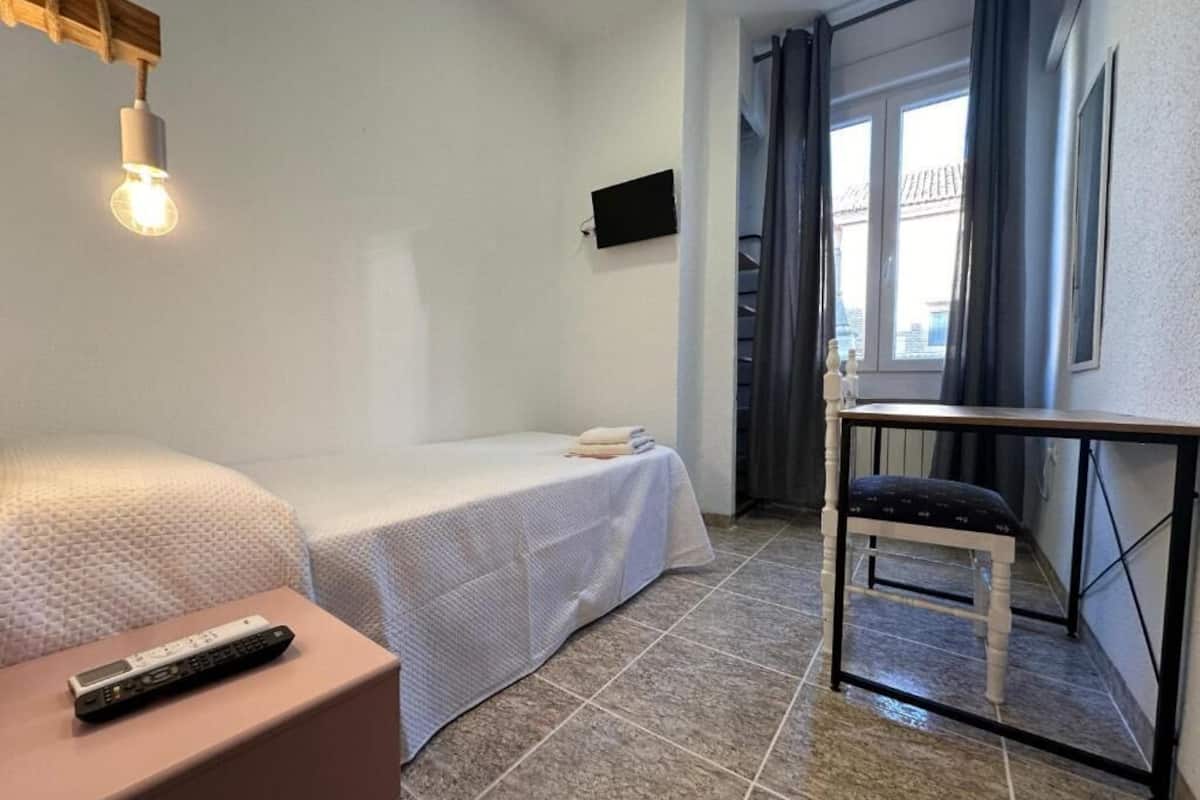 Quarto pequeno e bem organizado com uma cama, mesa de cabeceira com um controle remoto, um banquinho pequeno e uma janela com cortinas.