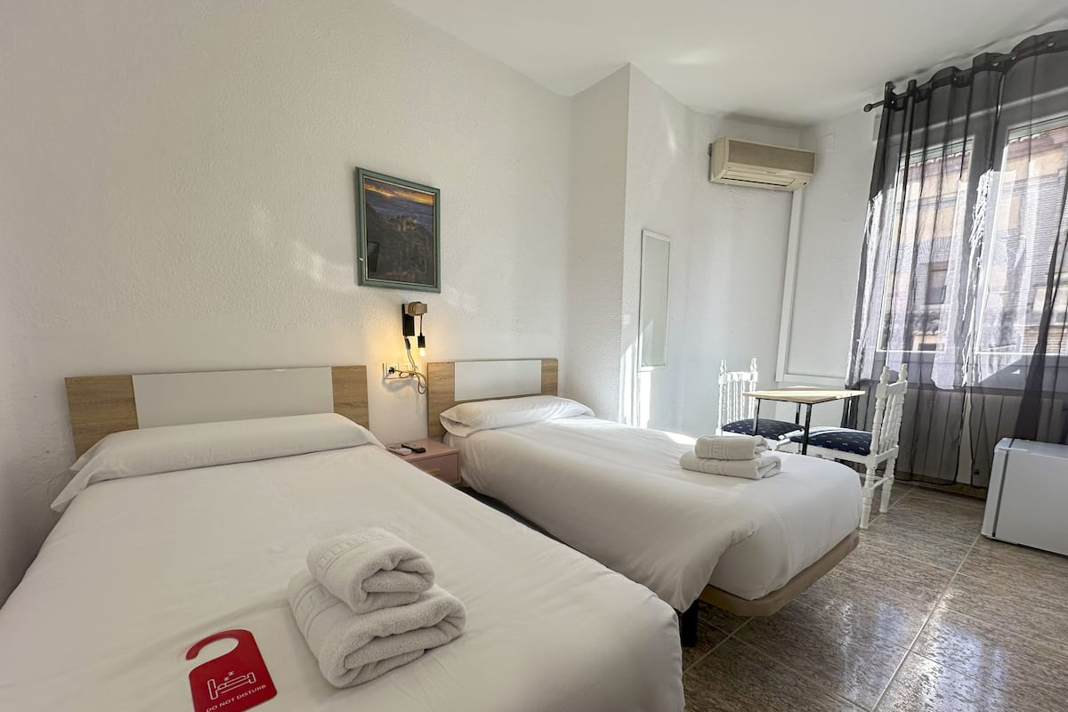 Quarto de hotel com duas camas, uma mesa pequena, uma cadeira, ar condicionado e um quadro na parede.