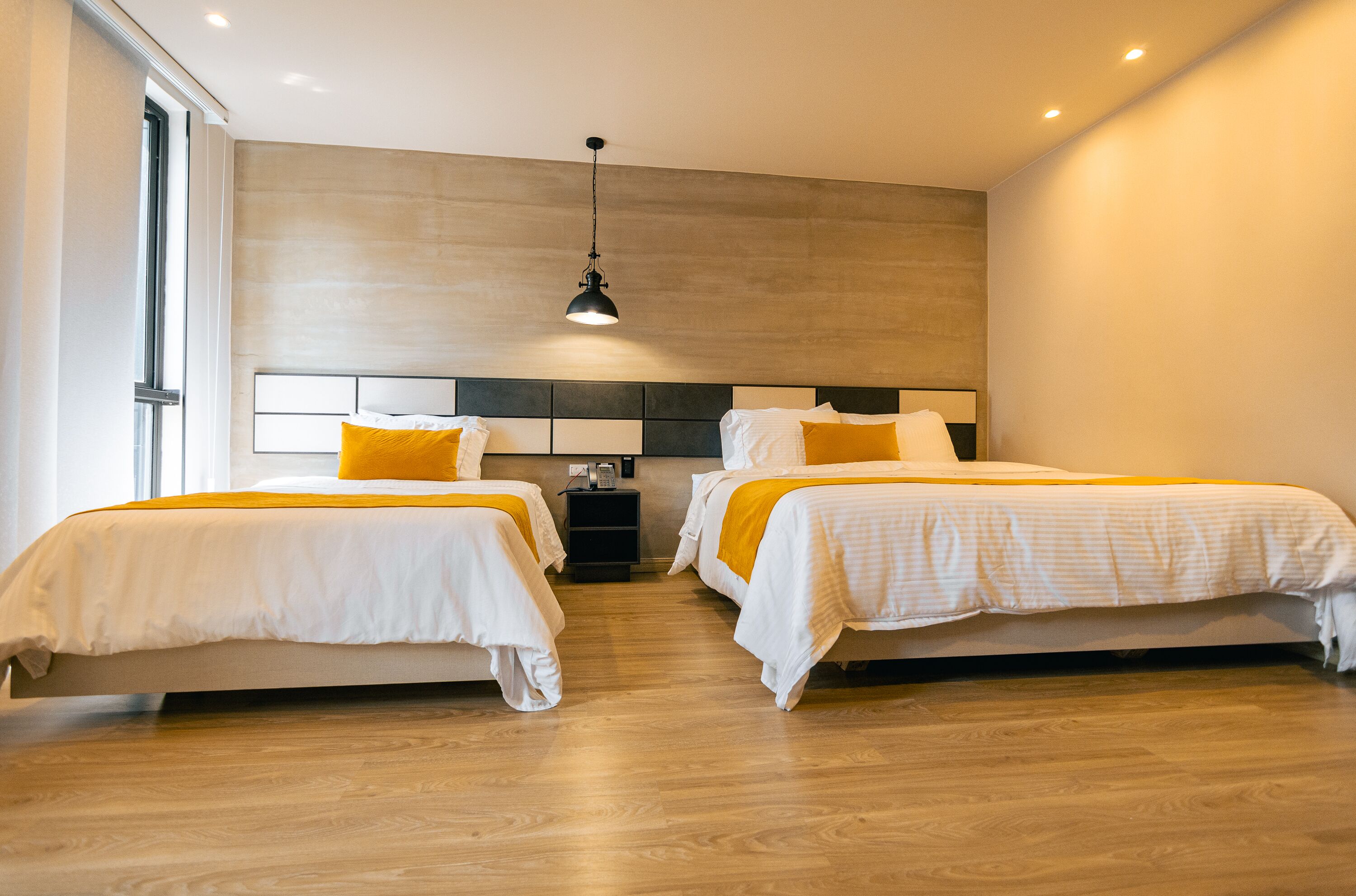 Quarto de hotel com duas camas, piso de madeira, cabeceira moderna e luminária pendente.