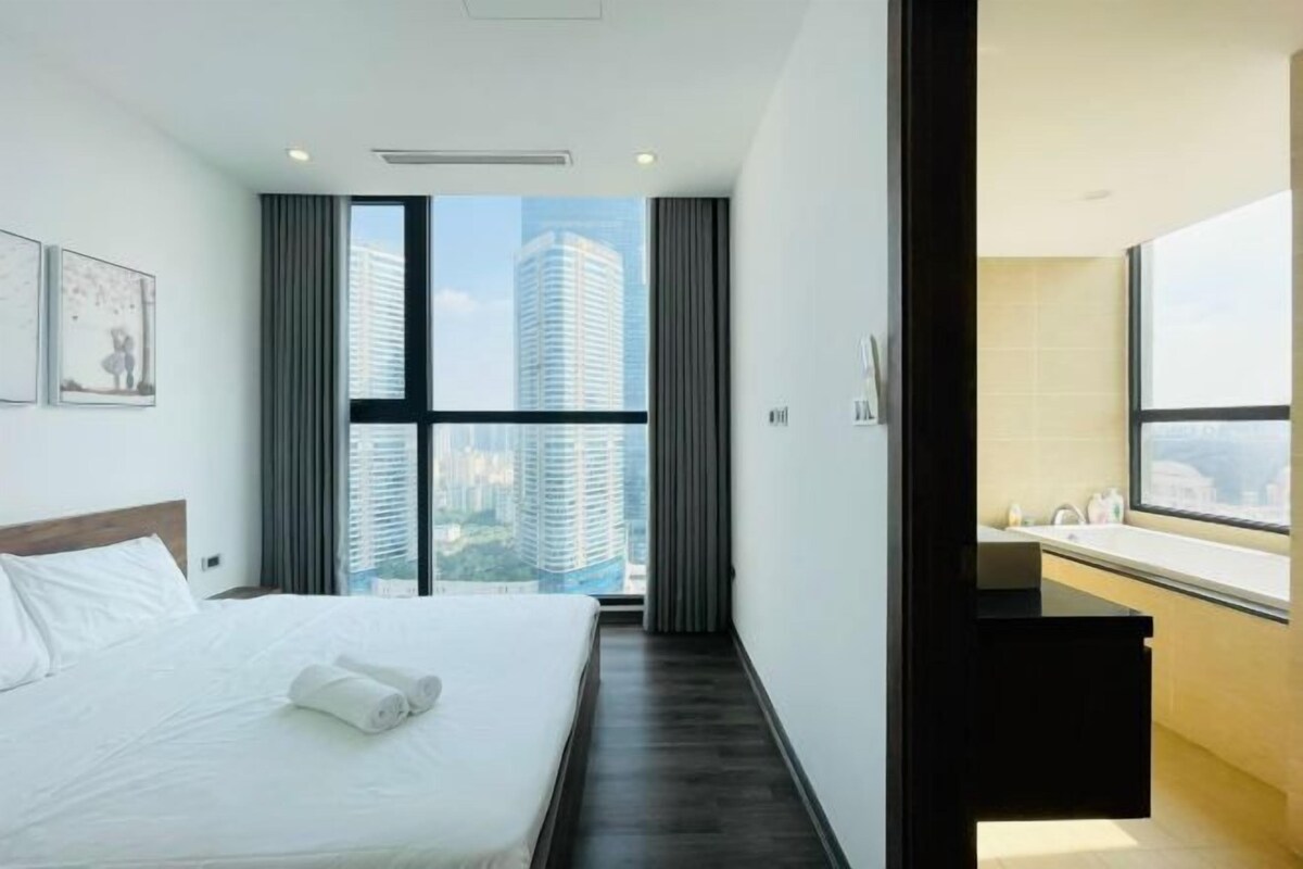 Quarto de hotel moderno com uma cama grande, vista para edifícios altos, banheiro com pia e espelho, e janela com cortinas.