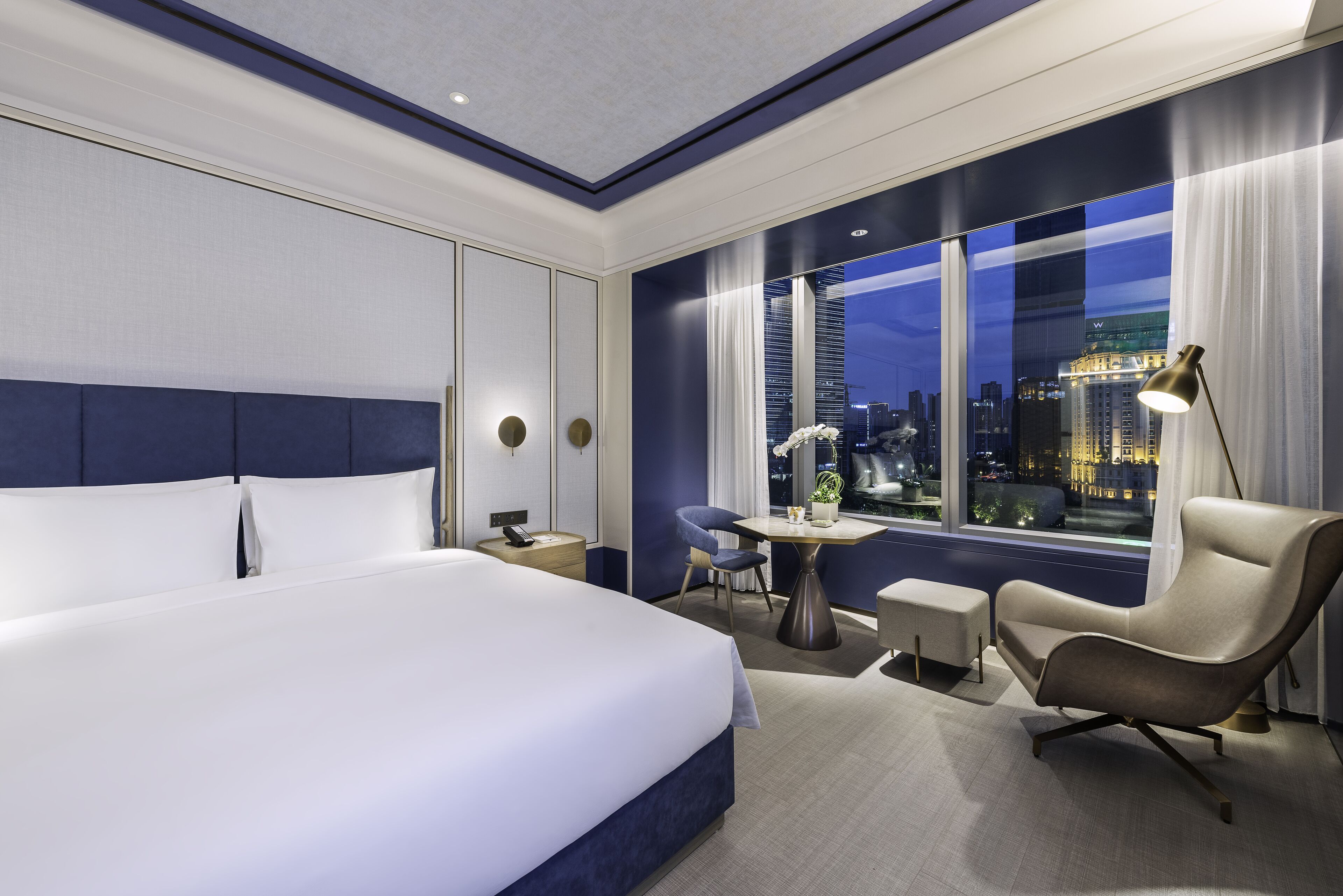Quarto de hotel moderno com uma cama grande, uma escrivaninha, uma cadeira e vista para a cidade à noite.