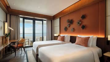 Quarto de hotel com duas camas, uma escrivaninha, uma cadeira e vista para o oceano.
