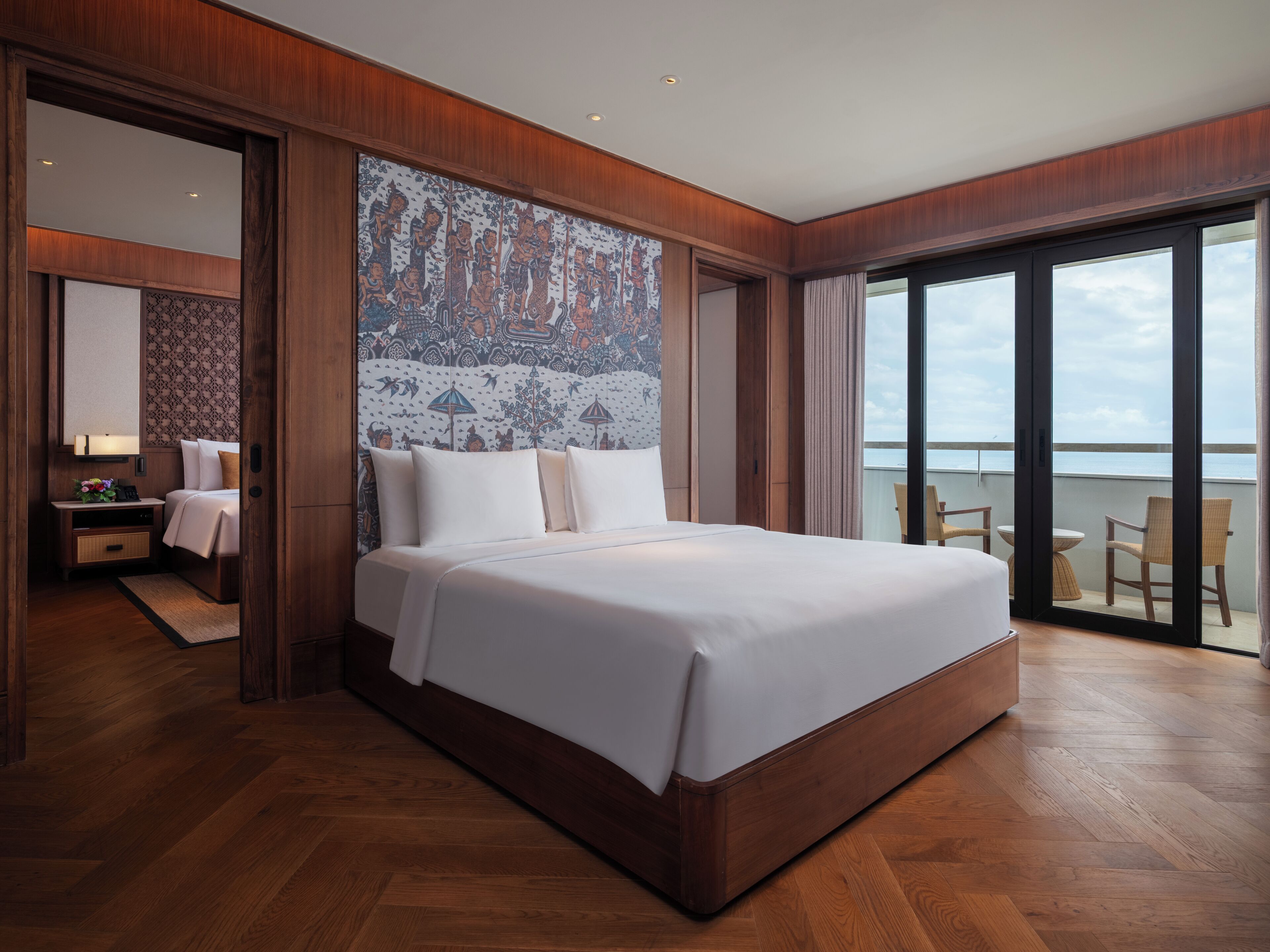 Quarto de hotel com uma cama grande, piso de madeira, varanda com vista e um detalhe decorativo na parede.