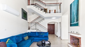 Uma sala de estar ampla com um sofá seccional azul, uma mesa de centro redonda e uma lareira. Uma escada com corrimão leva a um andar superior.